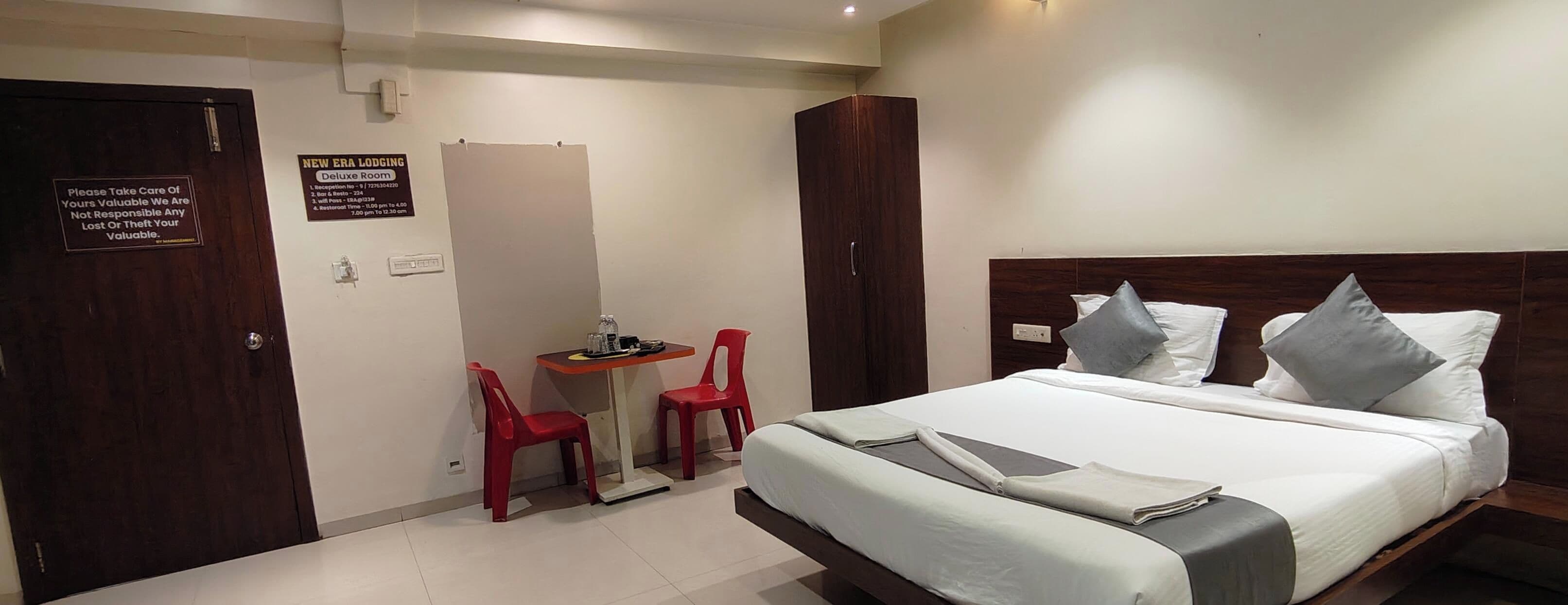 Quarto de hotel com cama, escrivaninha, duas cadeiras, guarda-roupa e um aviso na parede.