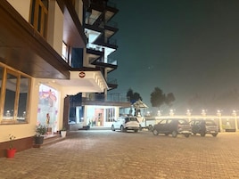 Edifício de vários andares com entrada no nível da rua, área de estacionamento com vários veículos e fachada bem iluminada.
