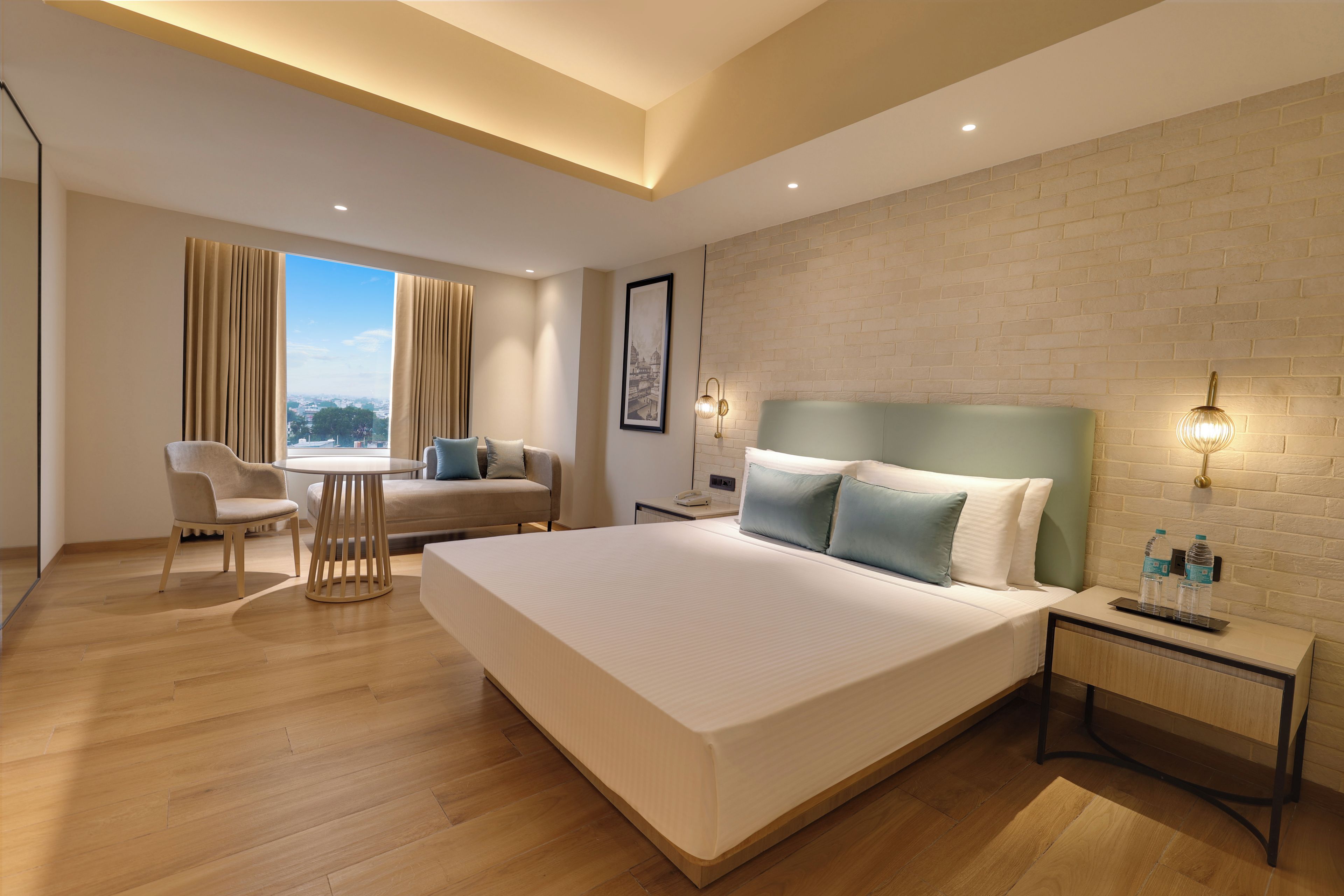 Quarto de hotel moderno com uma cama grande, área de estar com mesa e vista para a cidade.