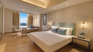 Quarto de hotel moderno com uma cama grande, área de estar com mesa e vista para a cidade.