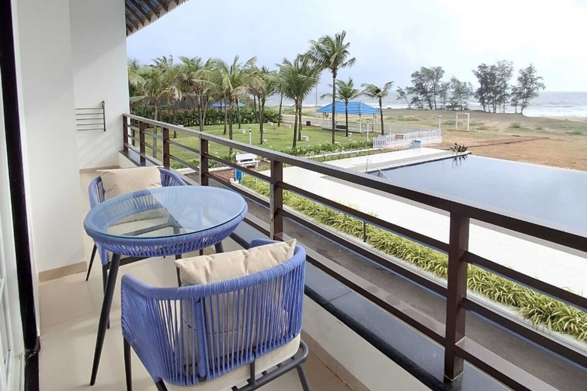 Varanda com mesa de vidro e cadeiras azuis, com vista para uma praia com palmeiras e céu claro.