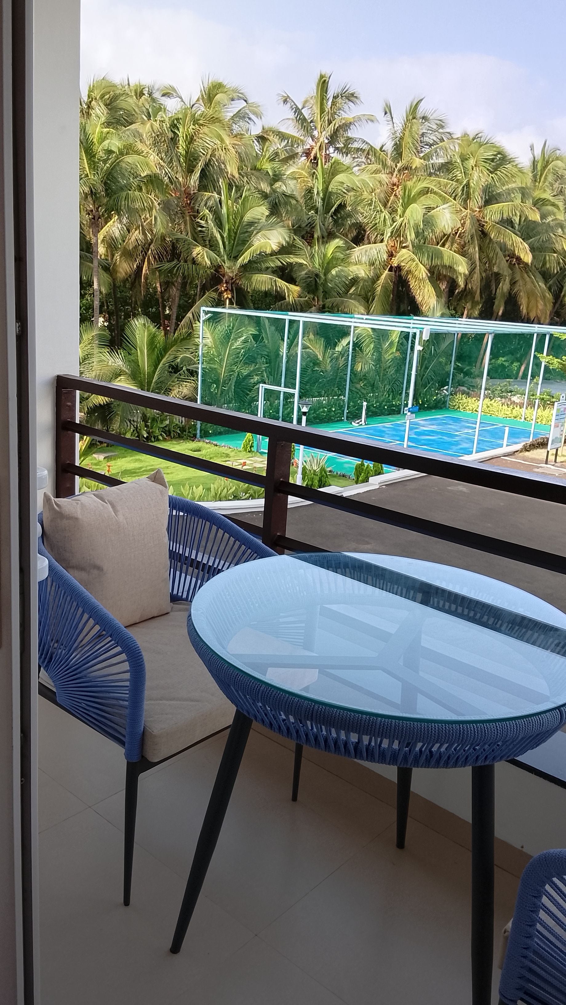 Varanda com mesa de vidro e cadeiras de vime azul, com vista para uma piscina e palmeiras.