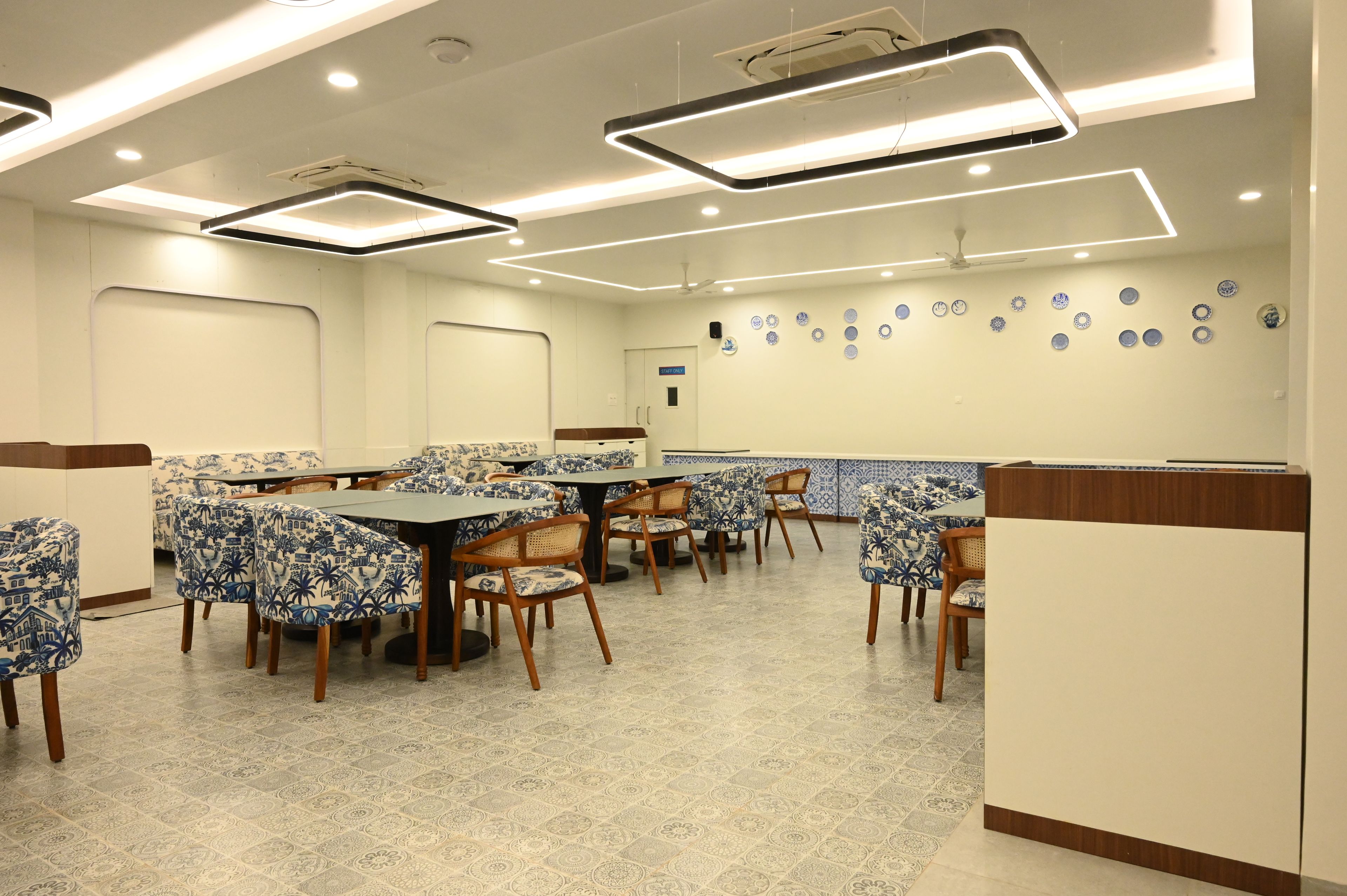 Um restaurante com várias mesas e cadeiras, teto com iluminação embutida e parede decorada com pratos.