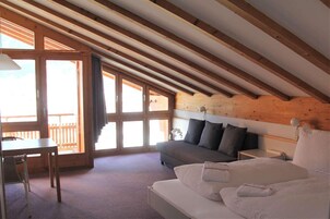 Quarto com teto inclinado, cama, sofá, escrivaninha e grandes janelas.