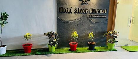 Uma área de recepção com placa indicando Hotel Silver Retreat by Tiliksha Hospitality, plantas em vasos e piso de azulejos.