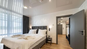 Quarto de hotel moderno com uma cama grande, mesa de cabeceira, guarda-roupa e vista pela janela com cortinas leves.