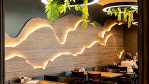 Interior de restaurante com mesas e cadeiras de madeira, parede com design de iluminação em forma de onda e plantas penduradas.