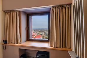 Quarto com janela que dá para uma vista urbana, uma escrivaninha com telefone e uma impressora preta no chão.