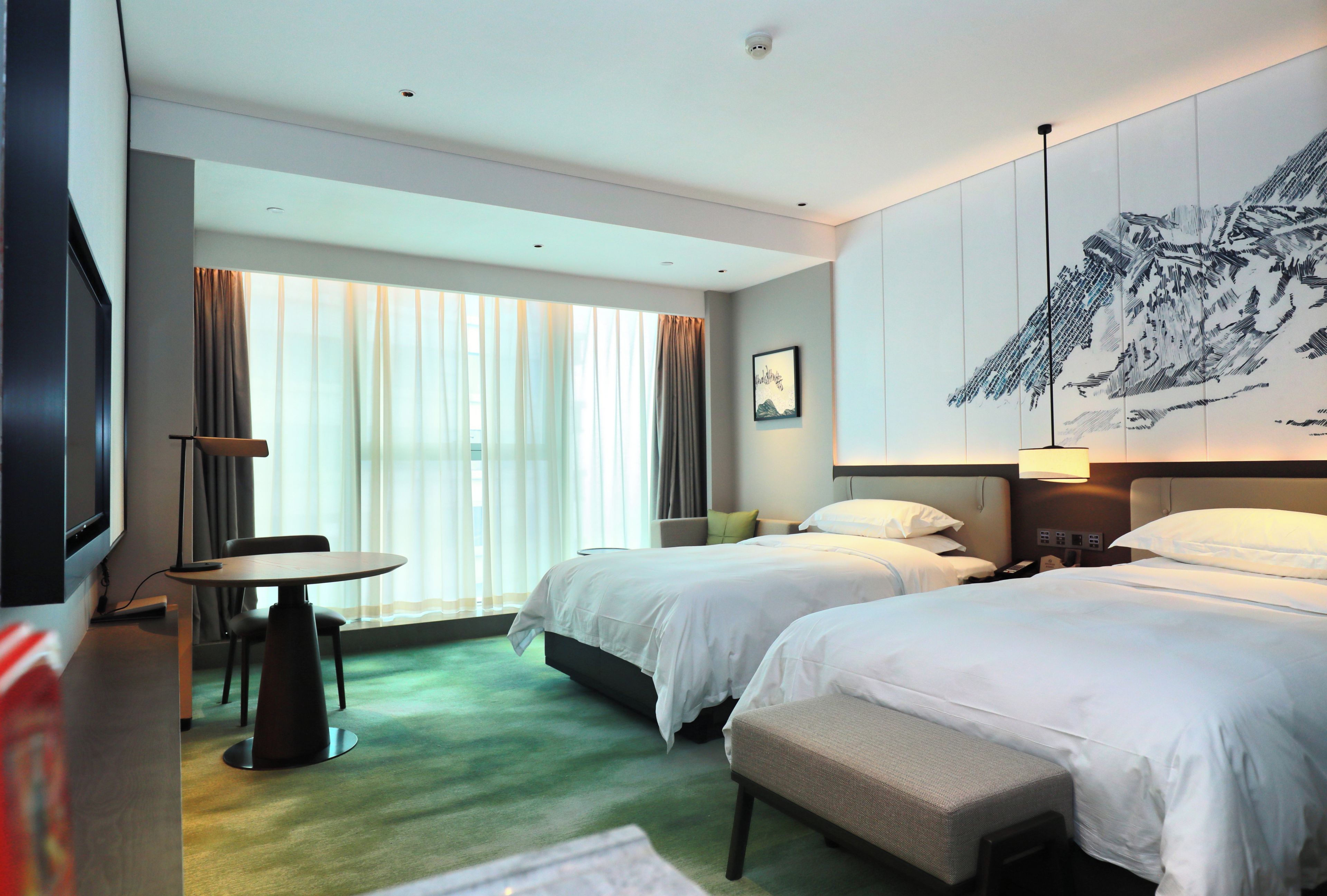Quarto de hotel com duas camas, uma escrivaninha, uma cadeira, uma televisão e um quadro grande na parede.