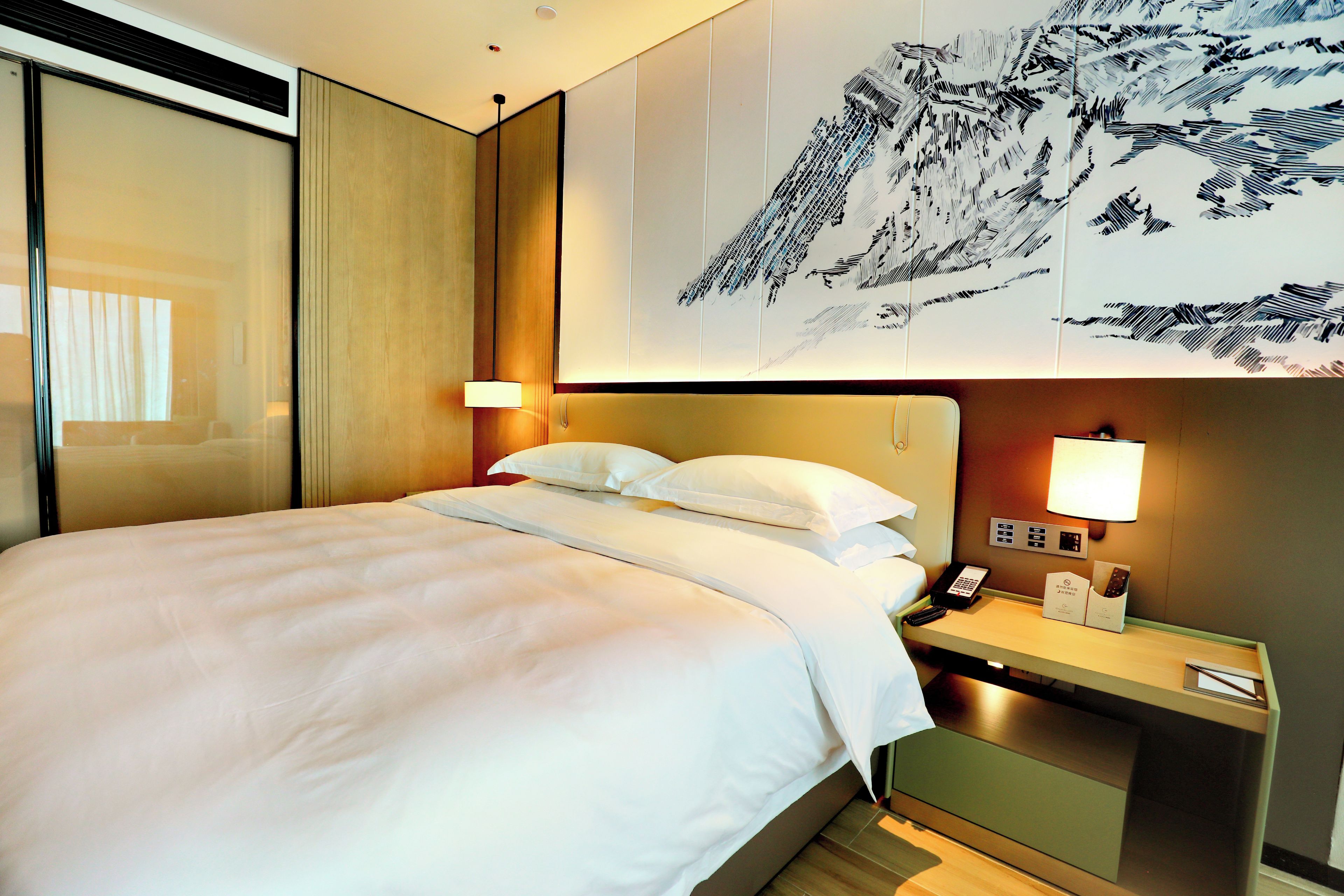 Quarto de hotel com cama, mesa de cabeceira, telefone e painel de controle fixado na parede.