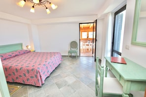Quarto com cama, escrivaninha, cadeira e vista para um pátio externo.