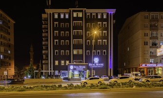 Hotel de vários andares com uma placa grande 'Real House', entrada bem iluminada e vista da rua com carros estacionados.