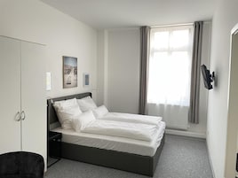 Quarto de hotel com cama, guarda-roupa, escrivaninha, cadeira, janela com cortinas e obras de arte na parede.