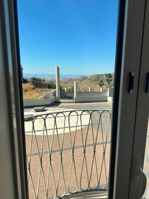 Vista pela janela, com grade de metal, céu claro e colinas ao longe.