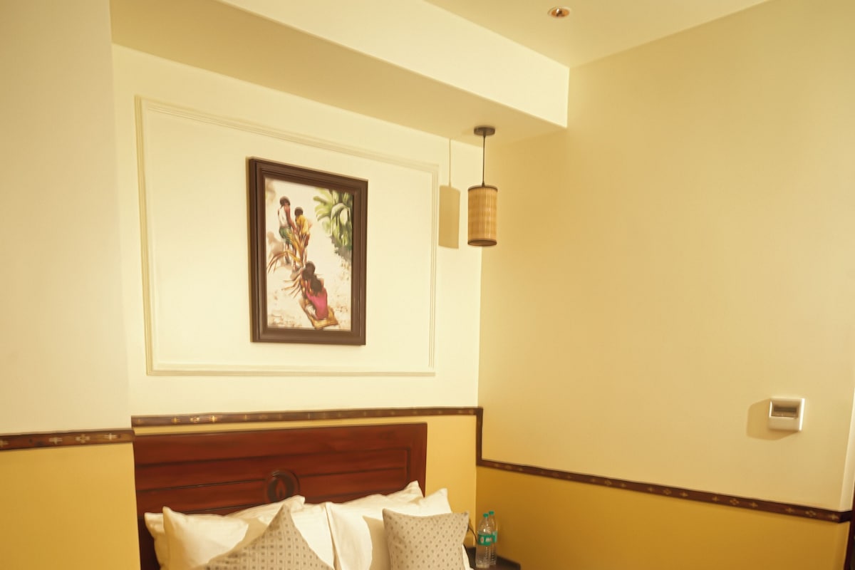 Quarto de hotel com cama, travesseiros, um quadro na parede e uma mesinha de cabeceira com uma garrafa.