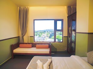 Quarto de hotel com cama, sofá, janela e cortinas, oferecendo vista para edifícios.