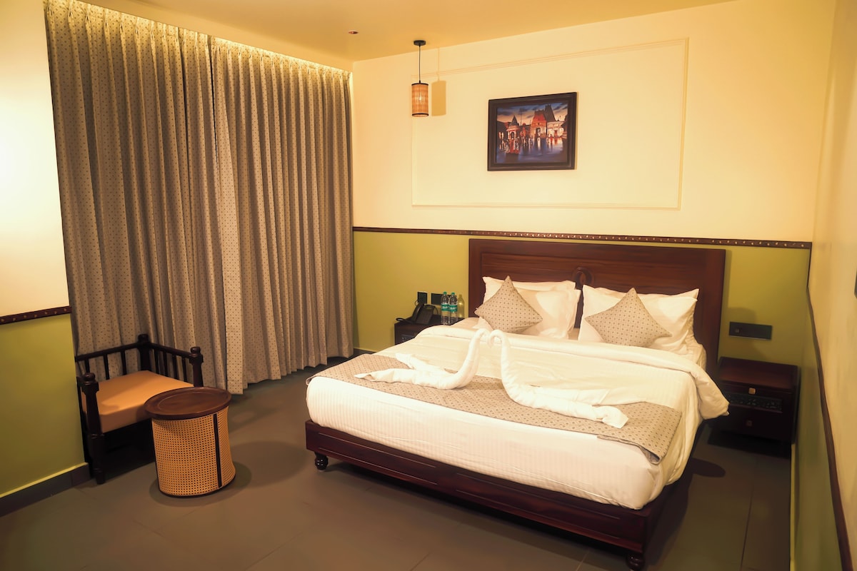 Quarto de hotel com cama grande, cabeceira de madeira, mesa redonda pequena, cesto de lixo e um quadro na parede.