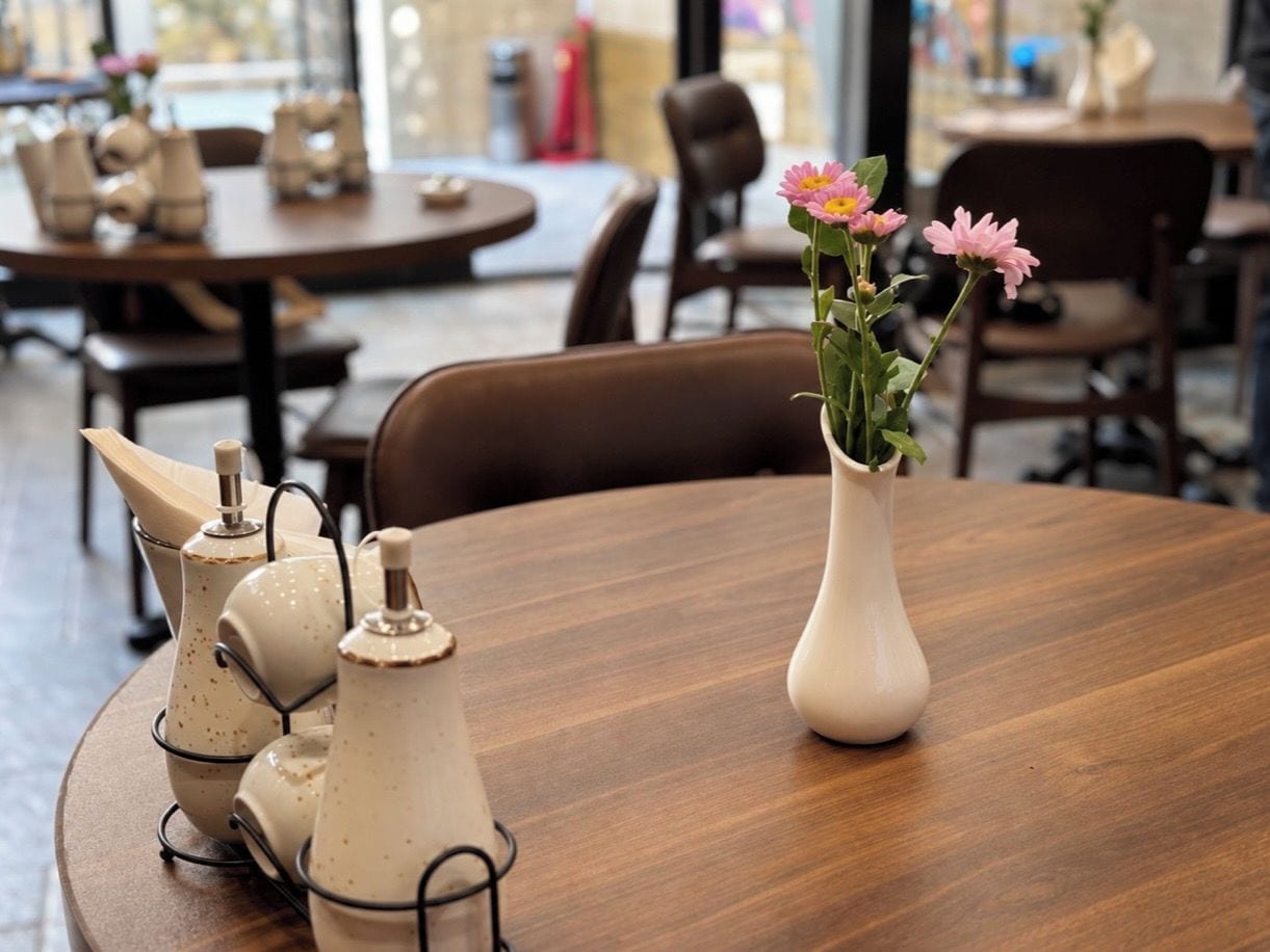 Uma sala de jantar com mesas redondas, cadeiras e um vaso com flores sobre uma das mesas.