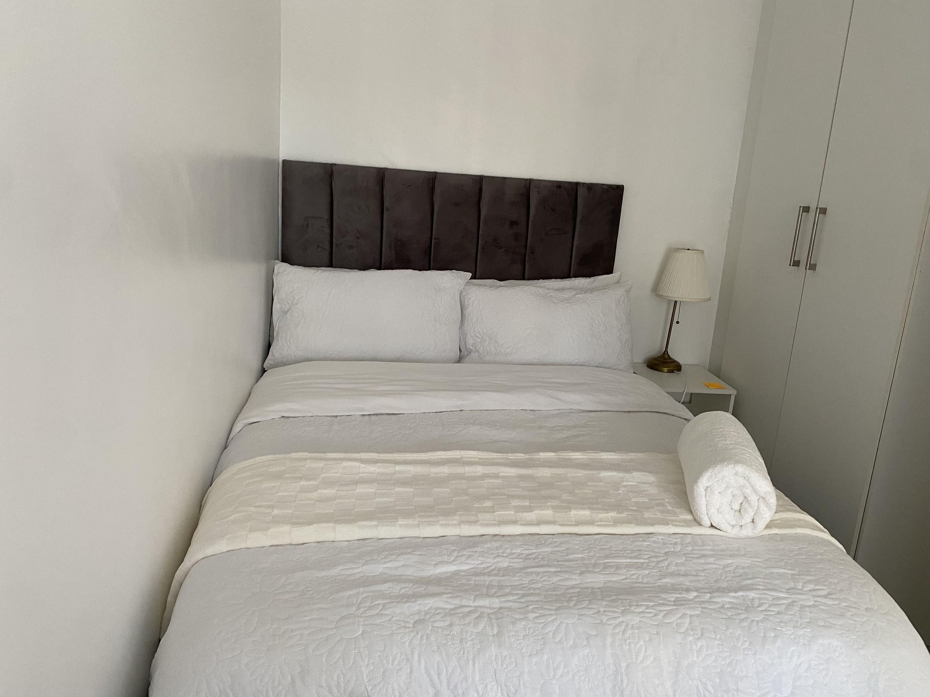 Cama bem arrumada com cabeceira, luminária de cabeceira e guarda-roupa.