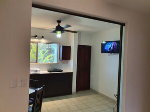 Cozinha compacta com área de refeições, ventilador de teto, TV fixada na parede e janela com persianas.