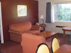 Quarto de hotel com sofá, mesa pequena, luminária e janela com cortinas.