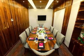 Uma sala de conferências com uma mesa de madeira comprida, cadeiras estofadas cinzentas, uma TV de tela plana na parede e uma planta decorativa no lado direito.