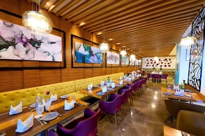 Um restaurante com painéis de madeira, um banco estofado amarelo, cadeiras roxas e obras de arte nas paredes.