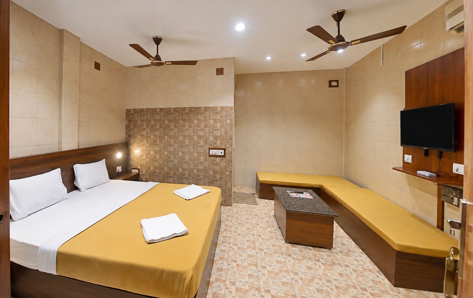 Quarto de hotel com cama, um banco, televisão e ventiladores de teto.