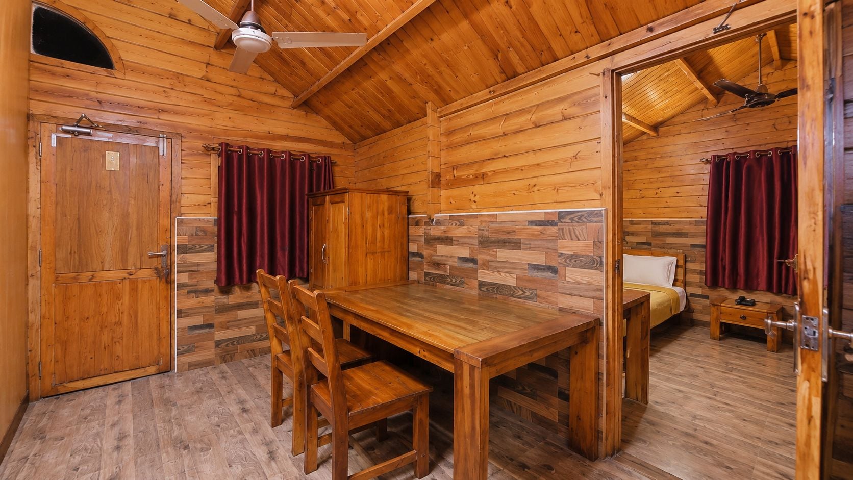 Interior de madeira com uma mesa de jantar, cadeiras, um armário e uma cama com cortina vermelha.