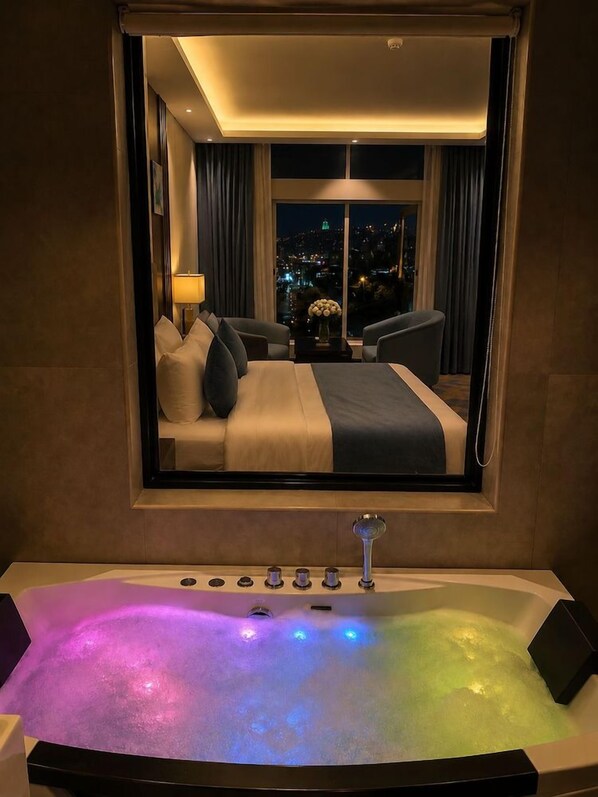 Quarto de hotel com um espelho grande refletindo uma cama, um banheiro com banheira e manípulos de controle, e vista para a cidade pela janela.