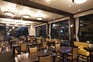 Espaço de refeições de hotel com várias mesas e cadeiras, grandes janelas e luminárias pendentes.