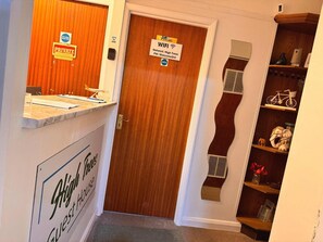 Uma área de recepção de hotel com uma porta de madeira, um balcão de recepção e uma placa na parede indicando 'High Trees Guest House'.