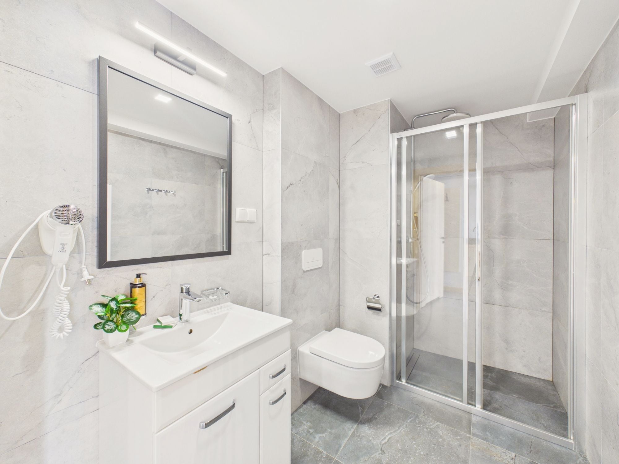 Casa de banho moderna com toucador branco, espelho grande, secador de cabelo, sanita e cabine de duche com parede de vidro.