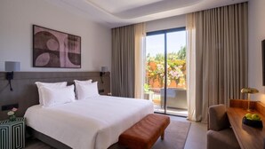 Quarto de hotel com uma cama grande, uma escrivaninha com abajur, um banco e vista para uma varanda com plantas.