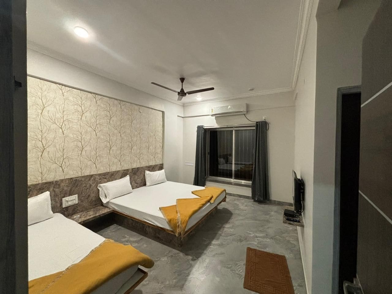 Quarto de hotel com duas camas, ventoinha no teto, aparelho de ar condicionado, televisão e janela com cortinas.