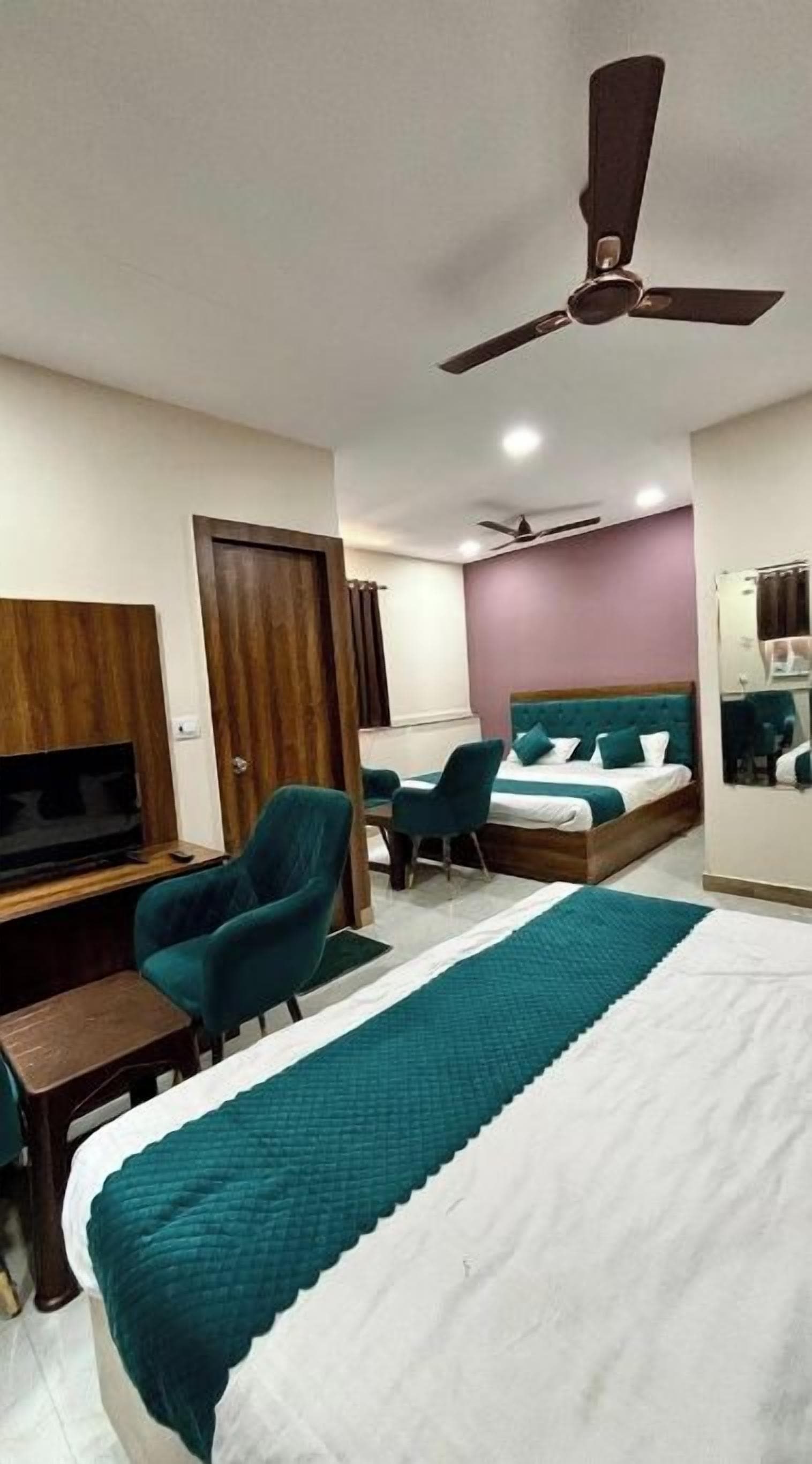 Quarto de hotel com uma cama, duas cadeiras, uma escrivaninha, televisão e ventoinha no teto.