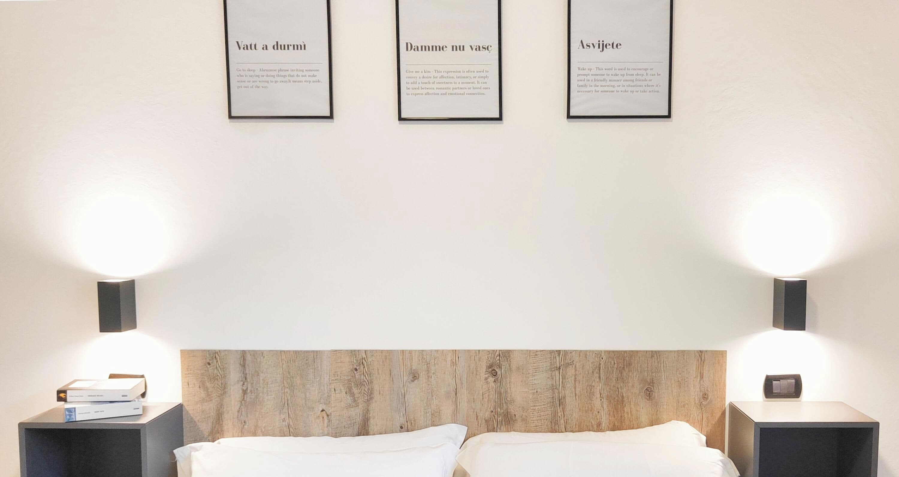 Quarto de hotel com cabeceira de madeira, duas luminárias de cabeceira, mesa de cabeceira com livros e três quadros na parede.