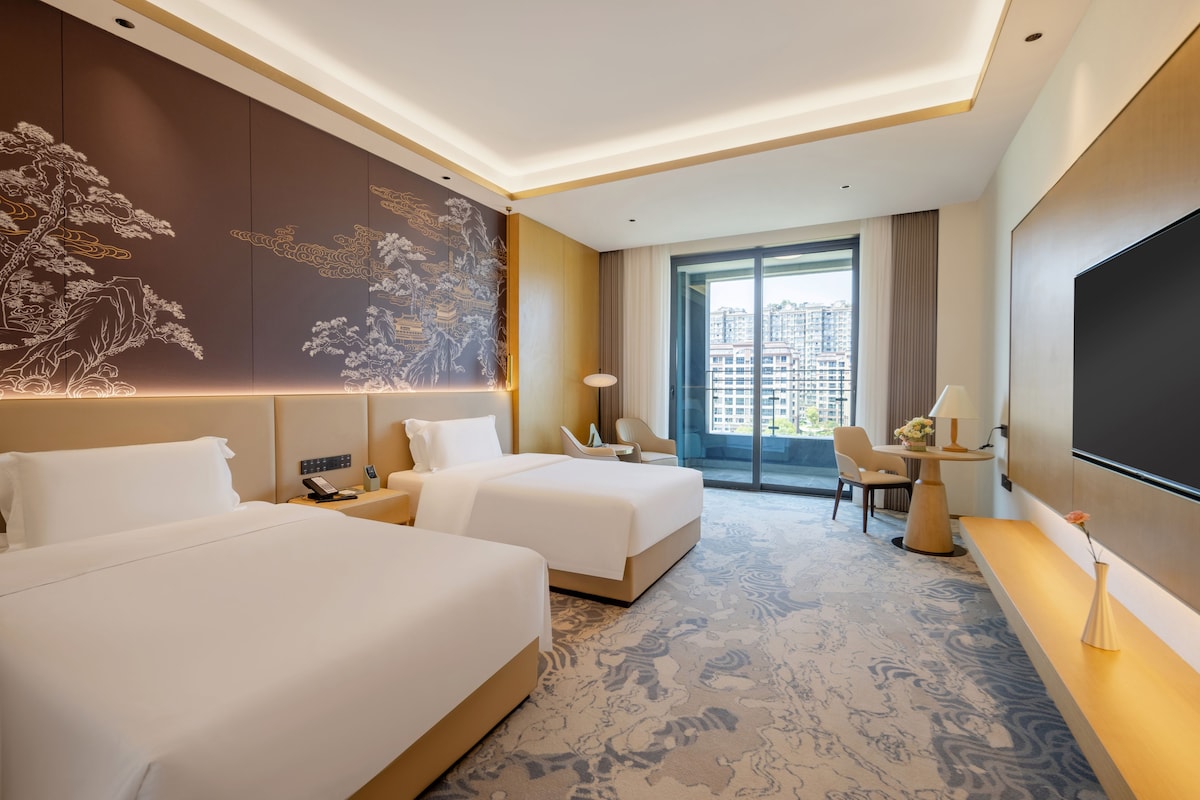 Quarto de hotel com duas camas, um mural grande na parede, uma varanda com vista para edifícios e uma TV de tela plana.