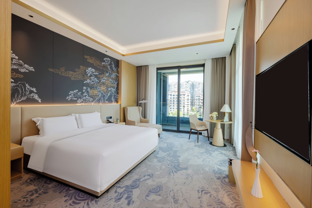 Quarto de hotel moderno com cama grande, televisão de tela plana, escrivaninha e vista para a paisagem urbana.