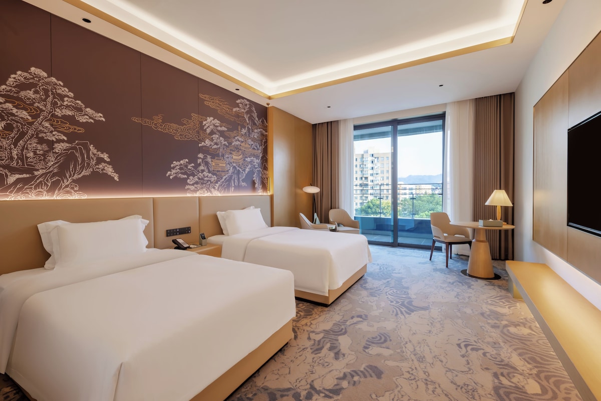 Quarto de hotel com duas camas, uma escrivaninha, uma cadeira, televisão e uma janela grande com vista para a cidade.