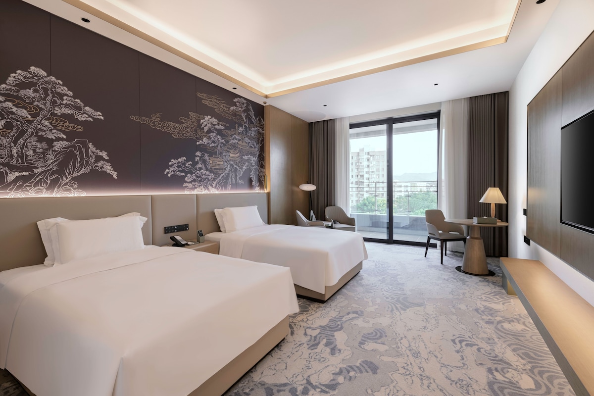 Quarto de hotel com duas camas, uma mesa, uma cadeira, televisão e uma janela grande com vista para edifícios.