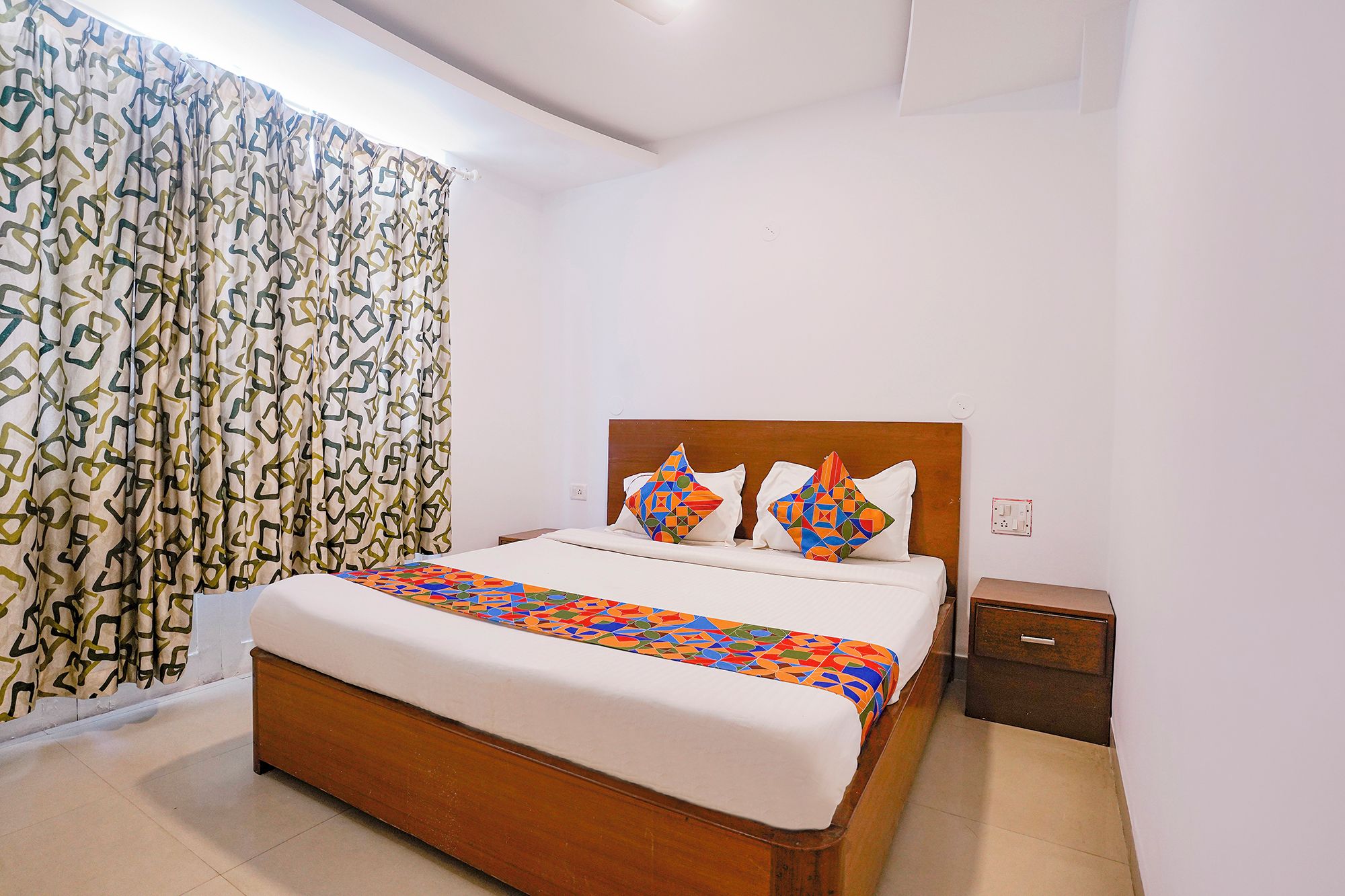 Uma cama bem arrumada com colcha estampada, duas almofadas com desenhos geométricos, cabeceira de madeira e uma pequena mesa de cabeceira com gaveta.
