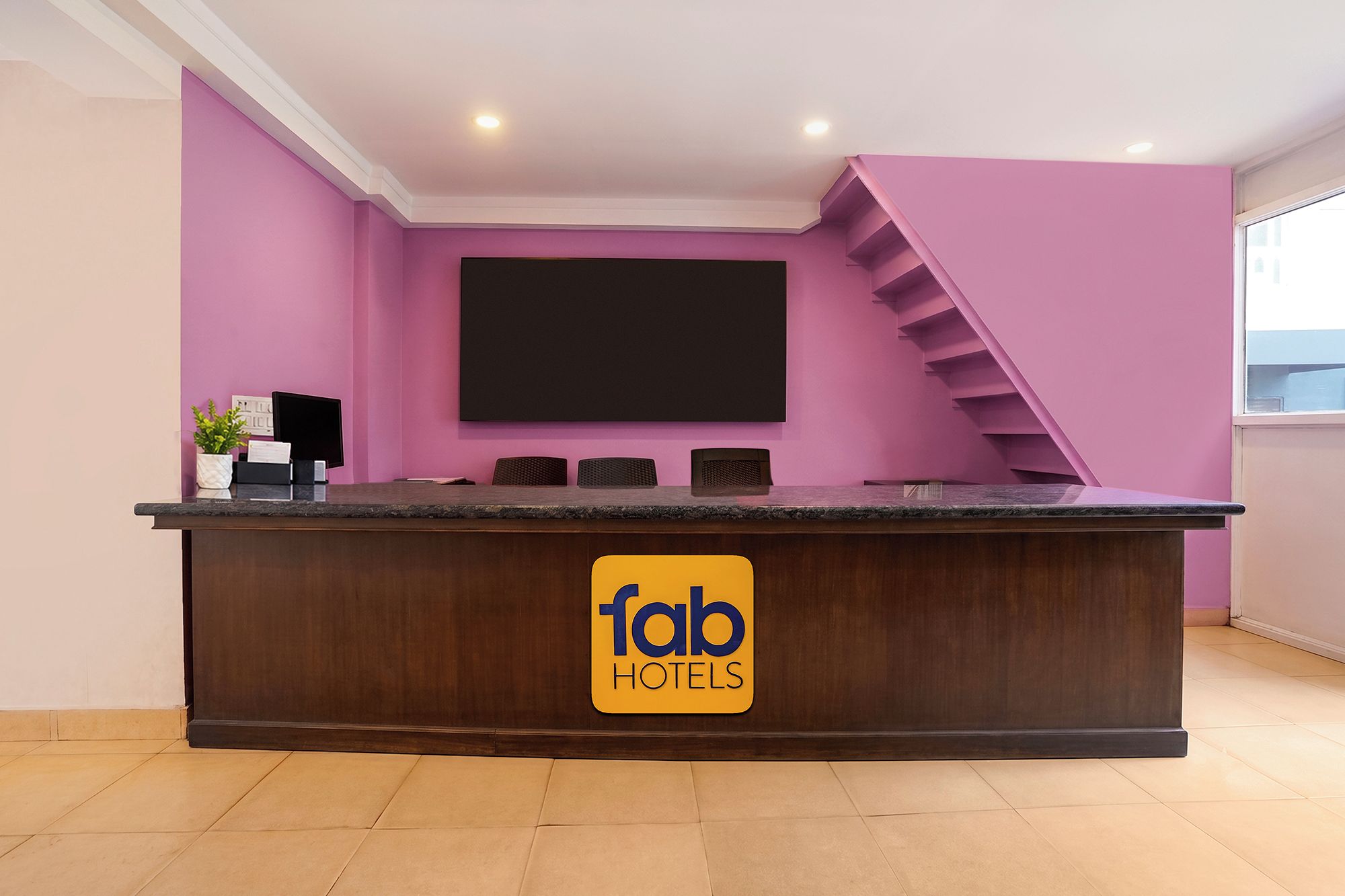 Uma mesa de recepção com a placa da Fab Hotels, uma televisão de tela plana na parede e uma escada que leva a um andar superior.