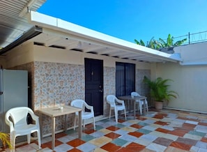 Um alpendre coberto com piso de azulejos, uma mesa com cadeiras, um vaso de planta e uma porta.