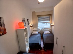 Quarto pequeno e compacto com duas camas de solteiro, um guarda-roupa branco, uma prateleira com um objeto vermelho e uma janela com persianas.