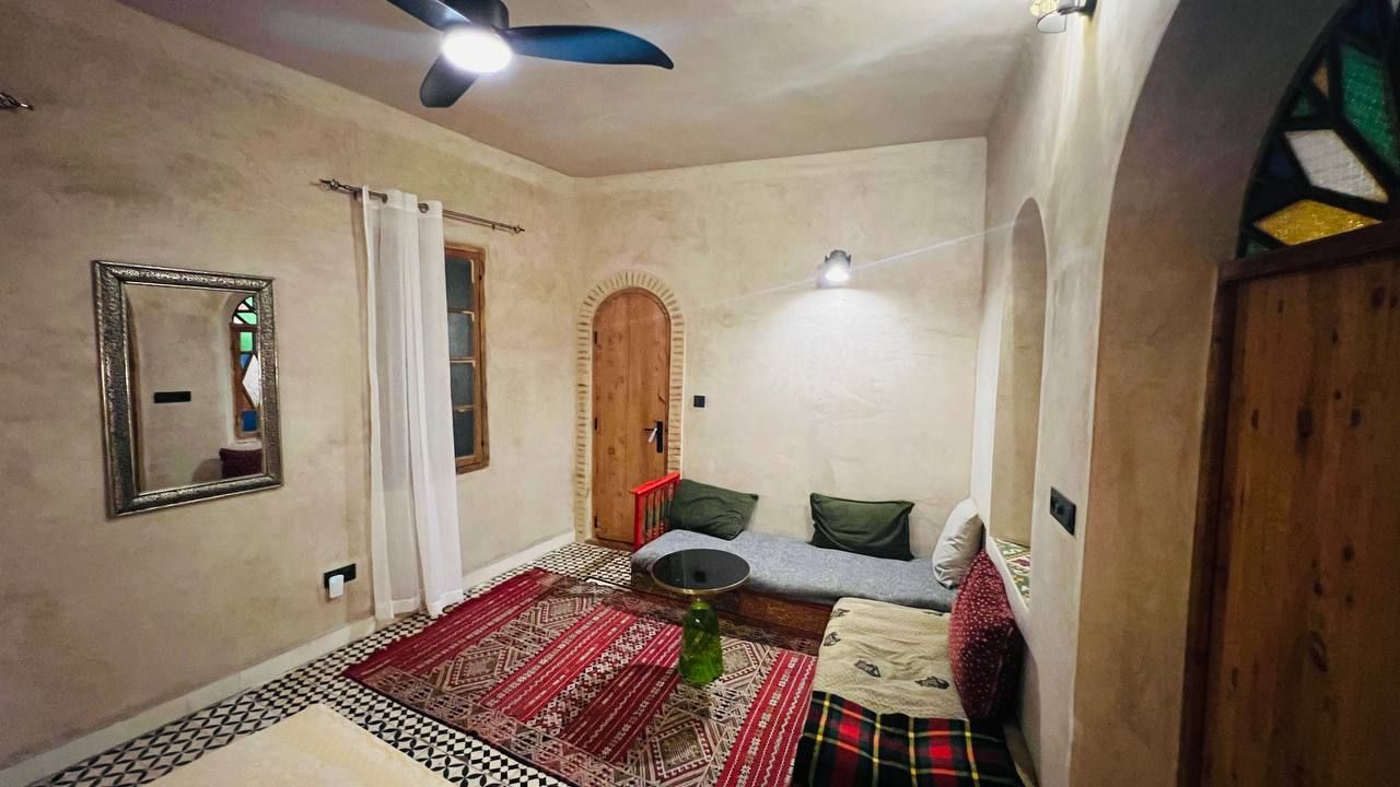 Um quarto com ventilador de teto, um sofá com almofadas, um tapete estampado, uma porta de madeira e um espelho na parede.