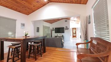 Uma sala de estar ampla com teto de madeira, uma mesa de jantar com banquetas, um sofá e uma televisão fixada na parede.