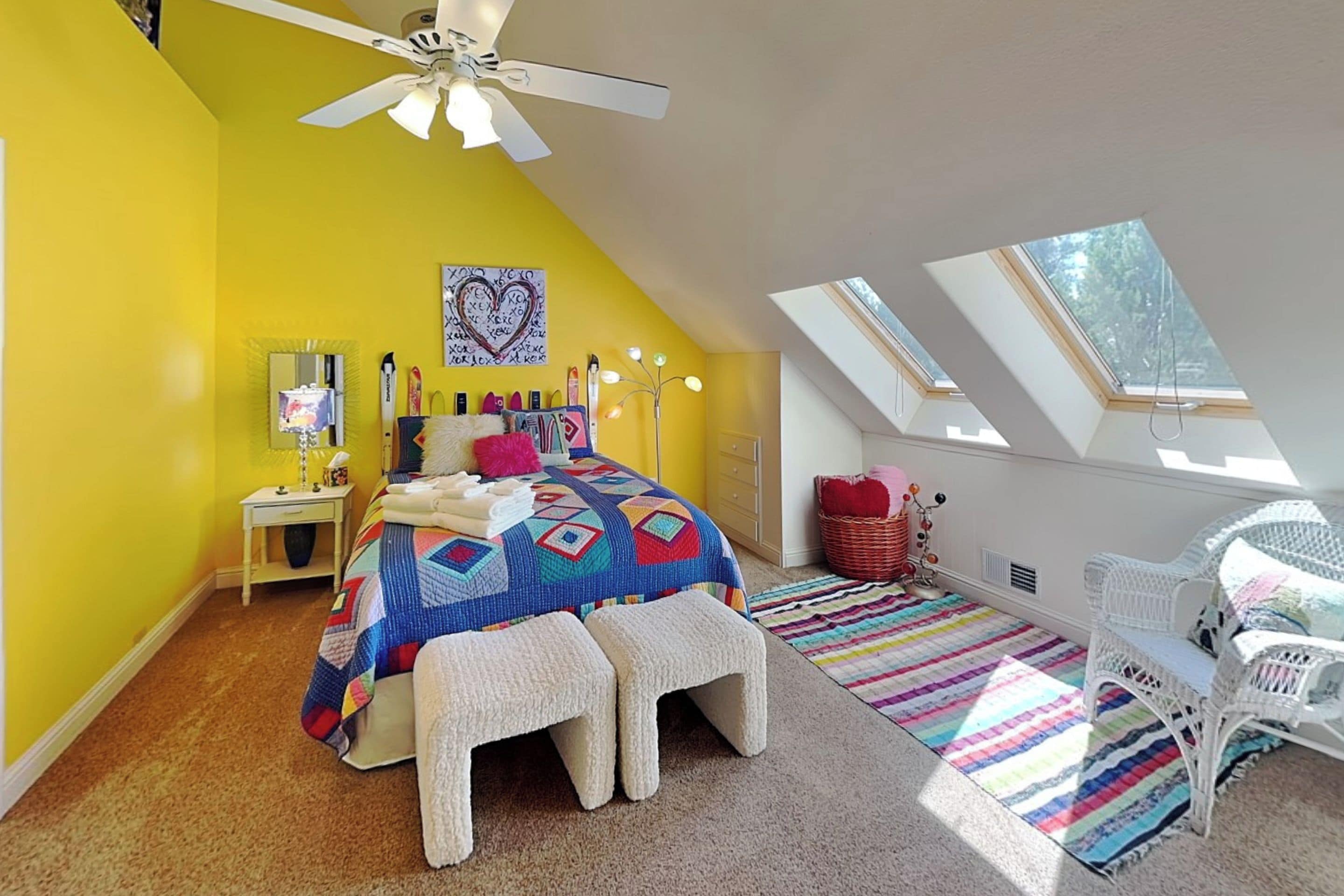 Quarto com teto inclinado, cama com colcha colorida, ventilador de teto, claraboia e um pequeno espaço de estar com uma cadeira e um tapete.
