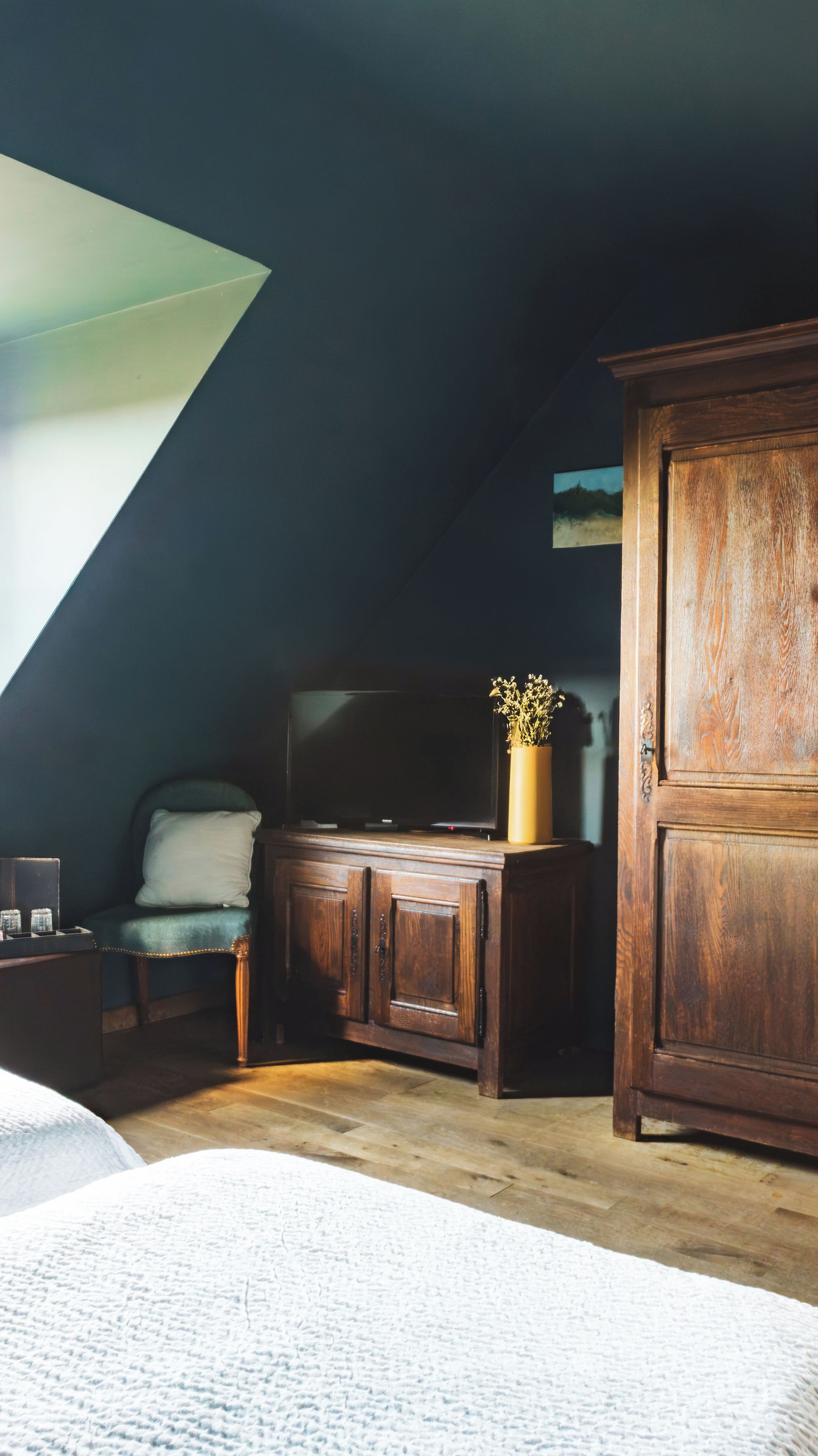 Quarto com teto inclinado, um armário de madeira, uma cadeira, uma televisão e um vaso com flores.