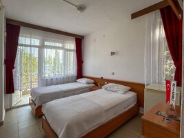 Quarto de hotel com duas camas de solteiro, cabeceira de madeira, mesa de cabeceira pequena com abajur, criado-mudo e uma janela com cortinas.